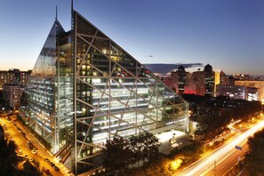 Um edifício moderno de vidro com design triangular, rodeado por uma paisagem urbana ao entardecer.