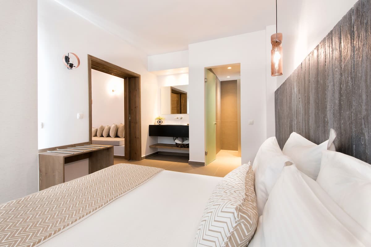 Quarto de hotel moderno com uma cama grande, um banco de madeira, um banco embutido com espelho, um pequeno banheiro com pia e espelho, e uma parede texturizada.