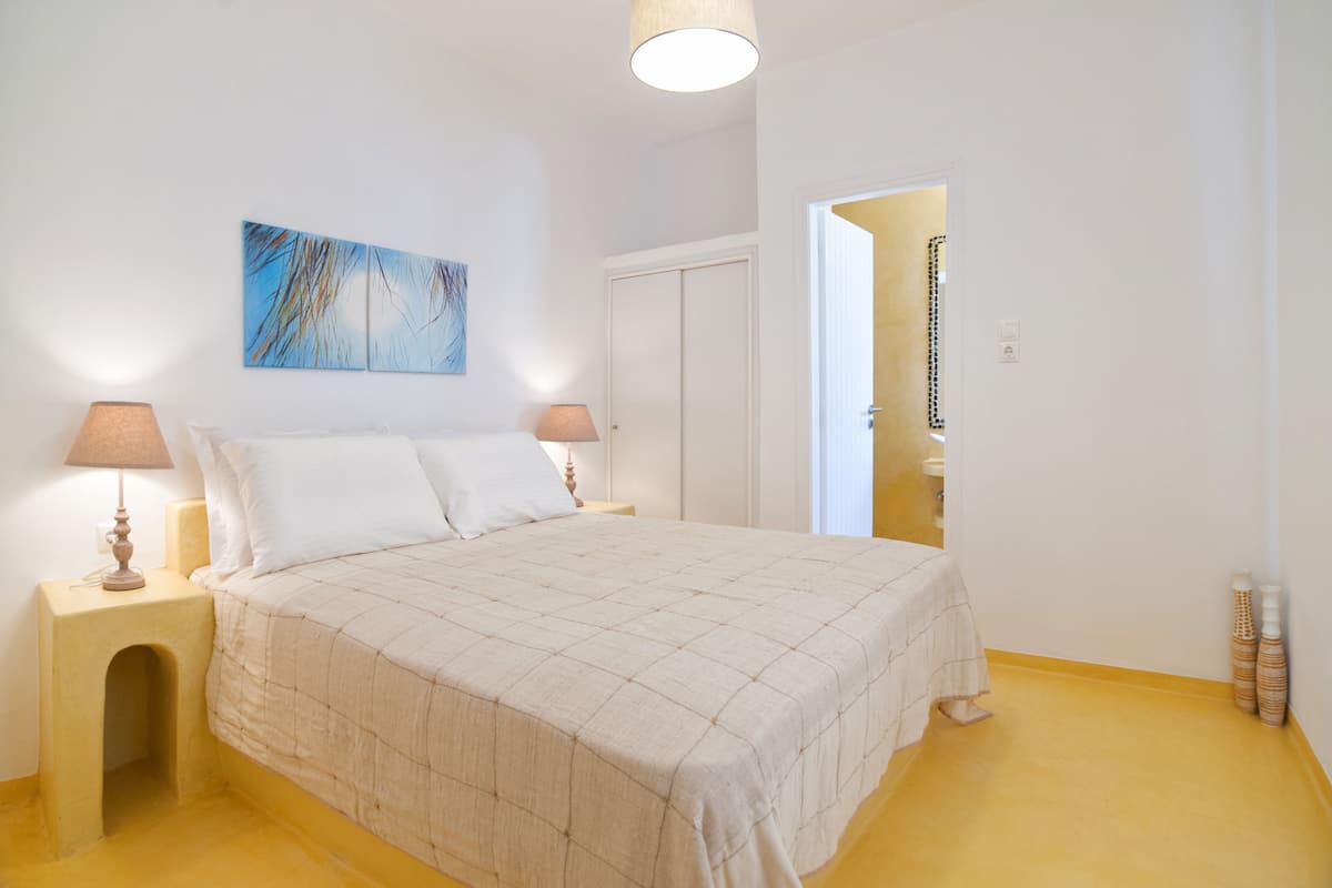 Quarto com cama, duas luminárias de cabeceira, um quadro na parede, uma porta e uma mesinha com vasos decorativos.