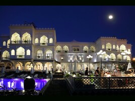 Um edifício tradicional com várias varandas e janelas arqueadas, área de piscina com guarda-sóis e céu noturno com a lua visível.