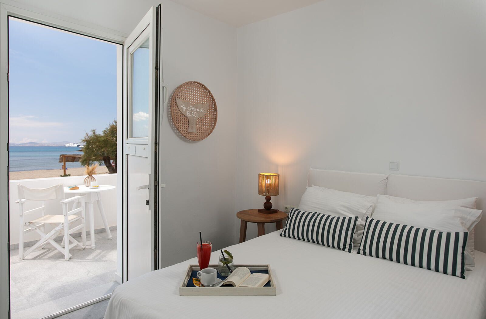 Quarto de hotel com cama, mesa de cabeceira com abajur, bandeja com bebida e um livro, vista para a praia e decoração de parede em vime.