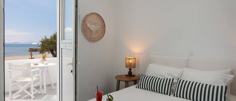 Quarto de hotel com cama, mesa de cabeceira com abajur, bandeja com bebida e um livro, vista para a praia e decoração de parede em vime.