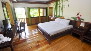 Quarto de hotel com cama de madeira, escrivaninha com televisão, cadeira e janela com cortinas.