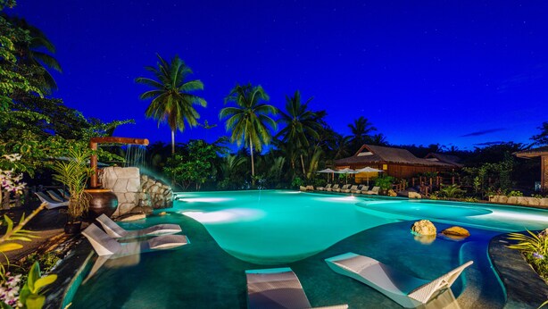 Piscina com espreguiçadeiras, palmeiras e palafitas de telhado de palha ao entardecer.