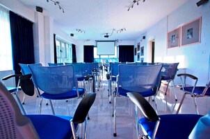 Uma sala de conferências com fileiras de cadeiras azuis, uma tela de projetor e uma grande janela.