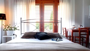 Quarto com cama, mesa de cabeceira com abajur, cadeira e vista para uma varanda com venezianas de madeira.