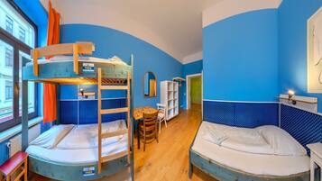 Quarto com beliche, escada de madeira, uma pequena mesa e uma cadeira. Há uma janela com cortinas laranja e vista para edifícios lá fora.