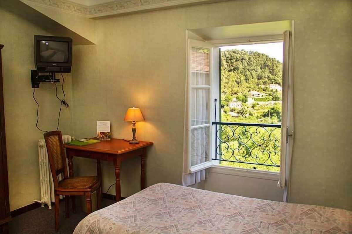 Quarto de hotel com cama, escrivaninha com luminária, cadeira, televisão na parede e janela com vista para uma paisagem verde.