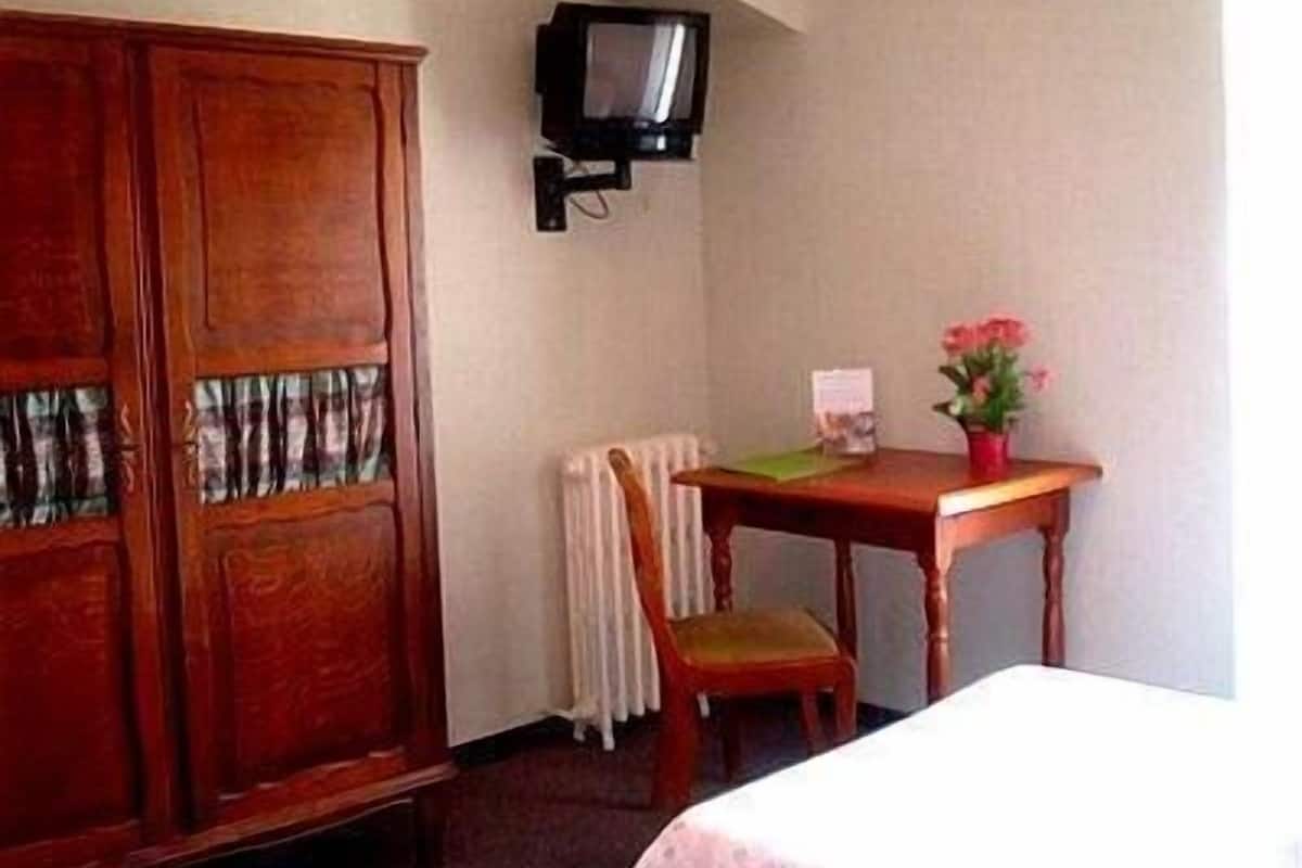 Quarto de hotel com guarda-roupa de madeira, uma pequena mesa com cadeira, radiador e televisão fixada na parede.