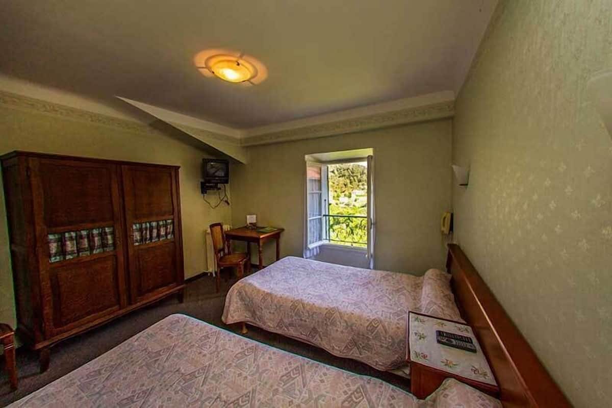 Quarto pequeno com duas camas, um armário de madeira, uma escrivaninha e uma janela com vista para o verde.