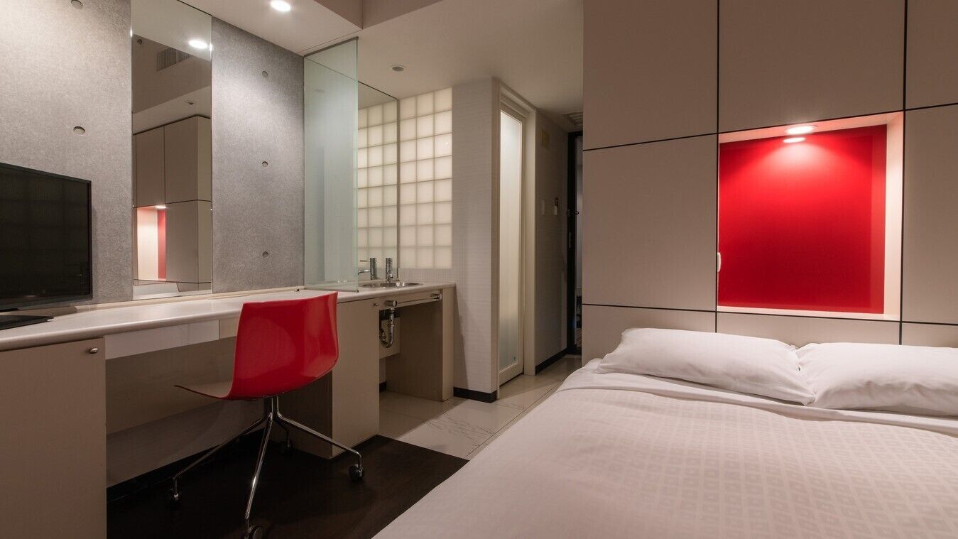 Quarto de hotel moderno com cama, escrivaninha com cadeira, espelho grande e parede com detalhe em vermelho.