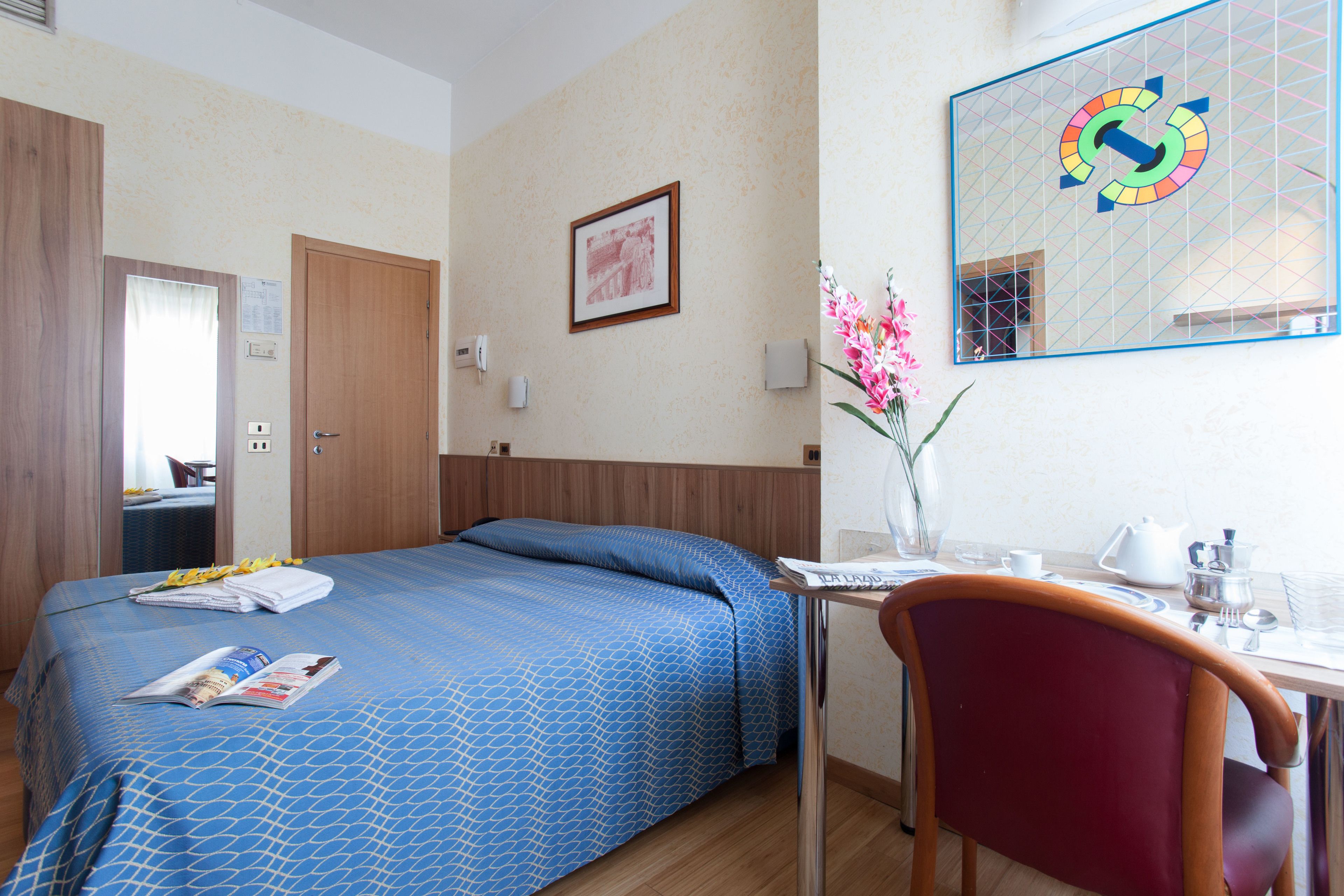 Quarto de hotel com cama, escrivaninha, cadeira, espelho e um vaso com flores.