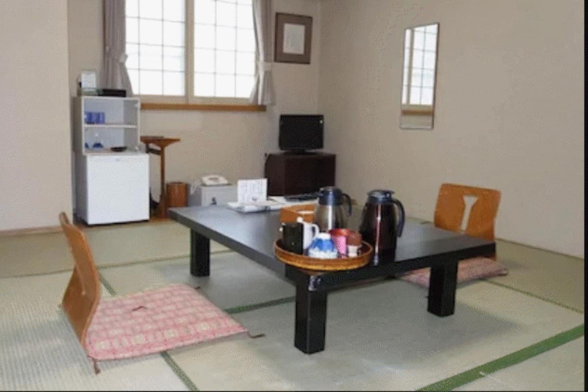 Quarto com piso de esteira de tatame, uma mesa baixa com uma bandeja de objetos, uma cadeira, uma pequena geladeira, uma televisão e uma janela com cortinas.