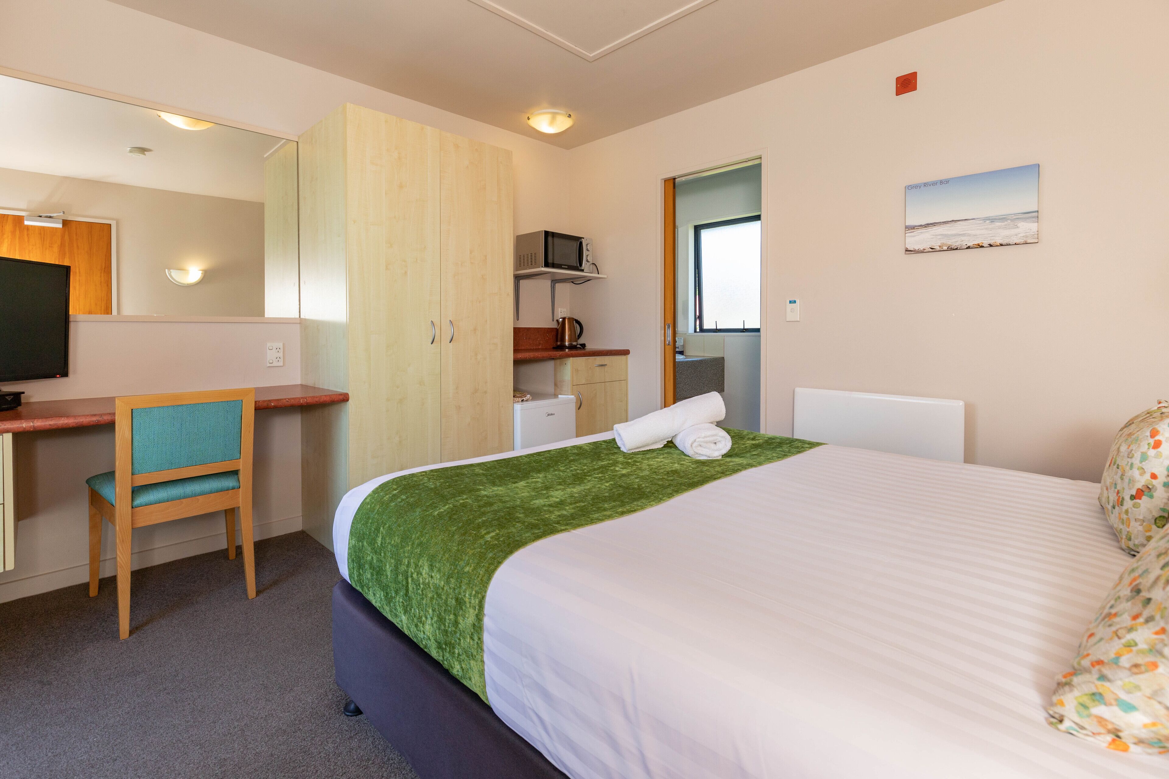 Quarto de hotel com cama, escrivaninha, cadeira, micro-ondas e um quadro na parede.