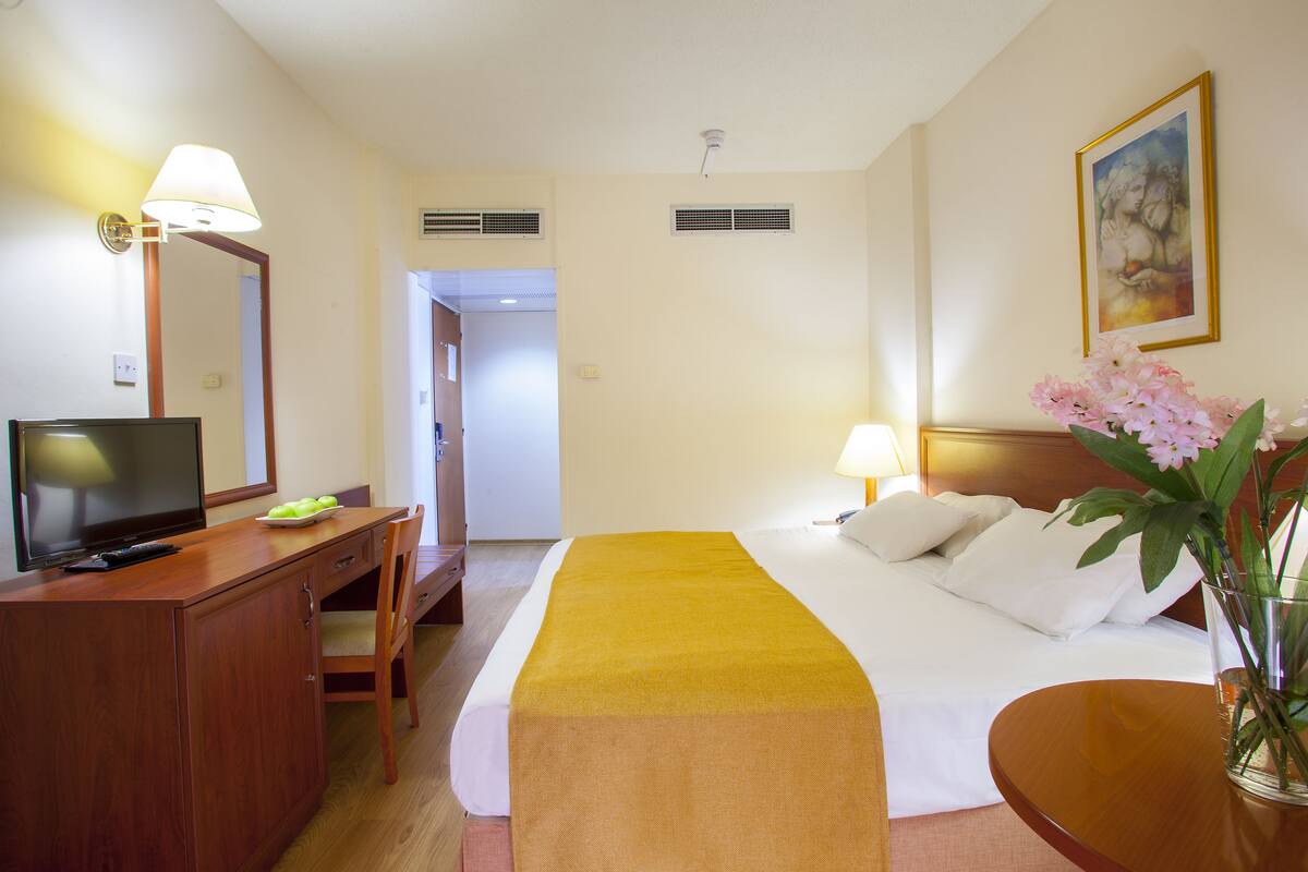 Quarto de hotel com cama, escrivaninha com televisão, cadeira, mesa de cabeceira com abajur e um vaso com flores.