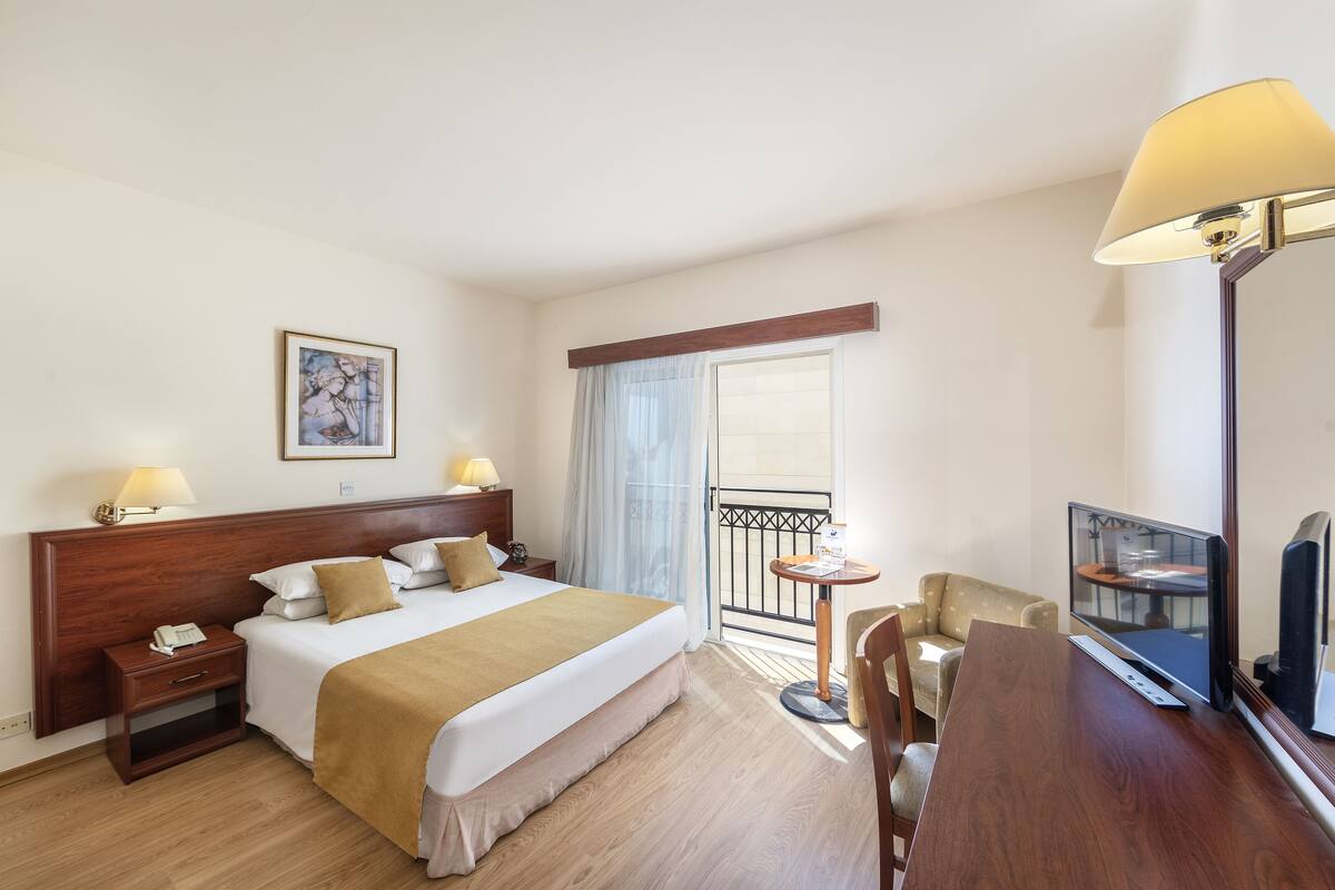 Quarto de hotel com uma cama grande, luminárias de cabeceira, escrivaninha com televisão, uma cadeira e varanda com vista.
