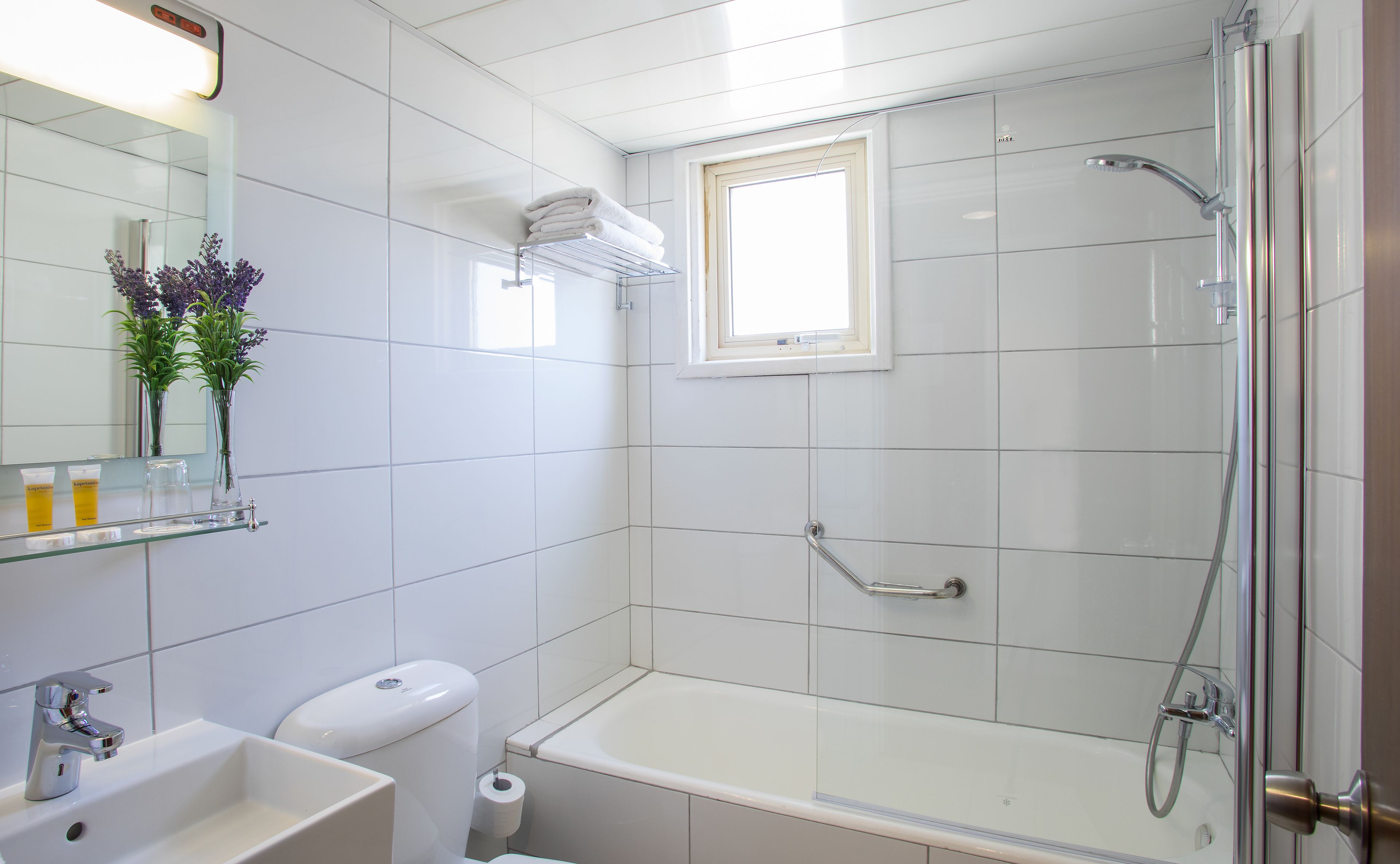 Casa de banho moderna com cabine de duche em vidro, banheira branca e sanita. Possui janela com luz natural e uma prateleira com produtos de higiene pessoal.