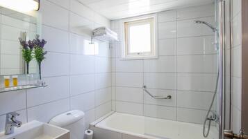Casa de banho moderna com cabine de duche em vidro, banheira branca e sanita. Possui janela com luz natural e uma prateleira com produtos de higiene pessoal.