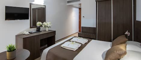 Quarto de hotel moderno com uma cama grande, mesa de cabeceira, escrivaninha, televisão de tela plana, espelho e guarda-roupa.