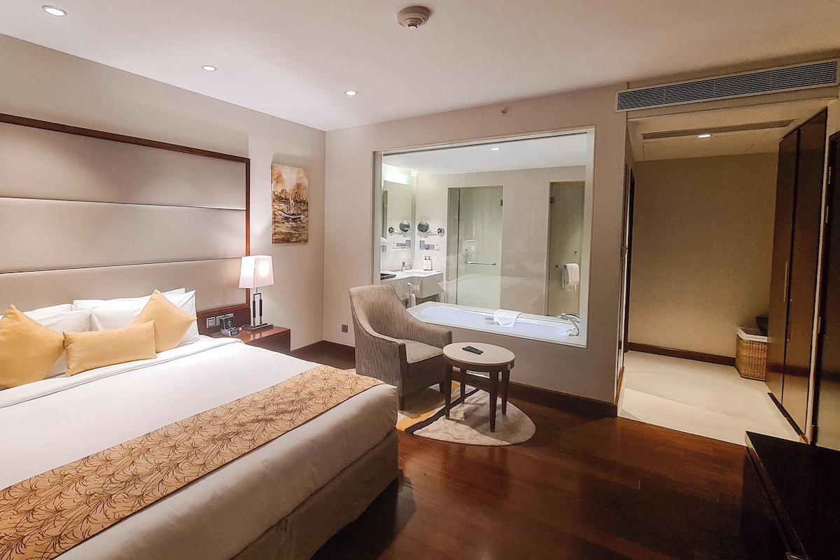 Quarto de hotel com uma cama grande, um espaço de estar com uma cadeira e uma pequena mesa, um espelho amplo e um banheiro visível através de uma porta de vidro.