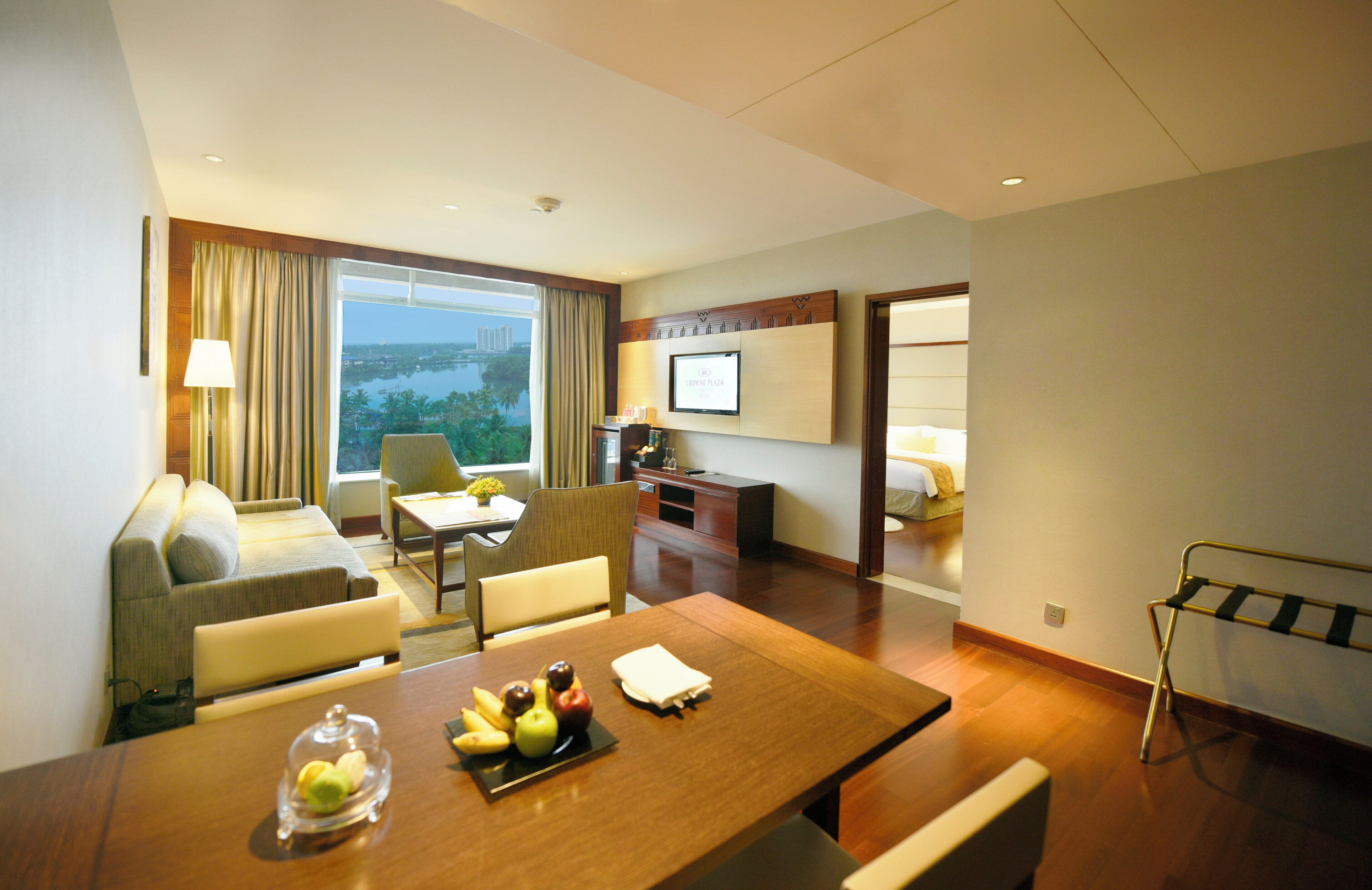 Quarto de hotel com uma mesa de jantar, cadeiras, um sofá, uma televisão e vista para a paisagem urbana.