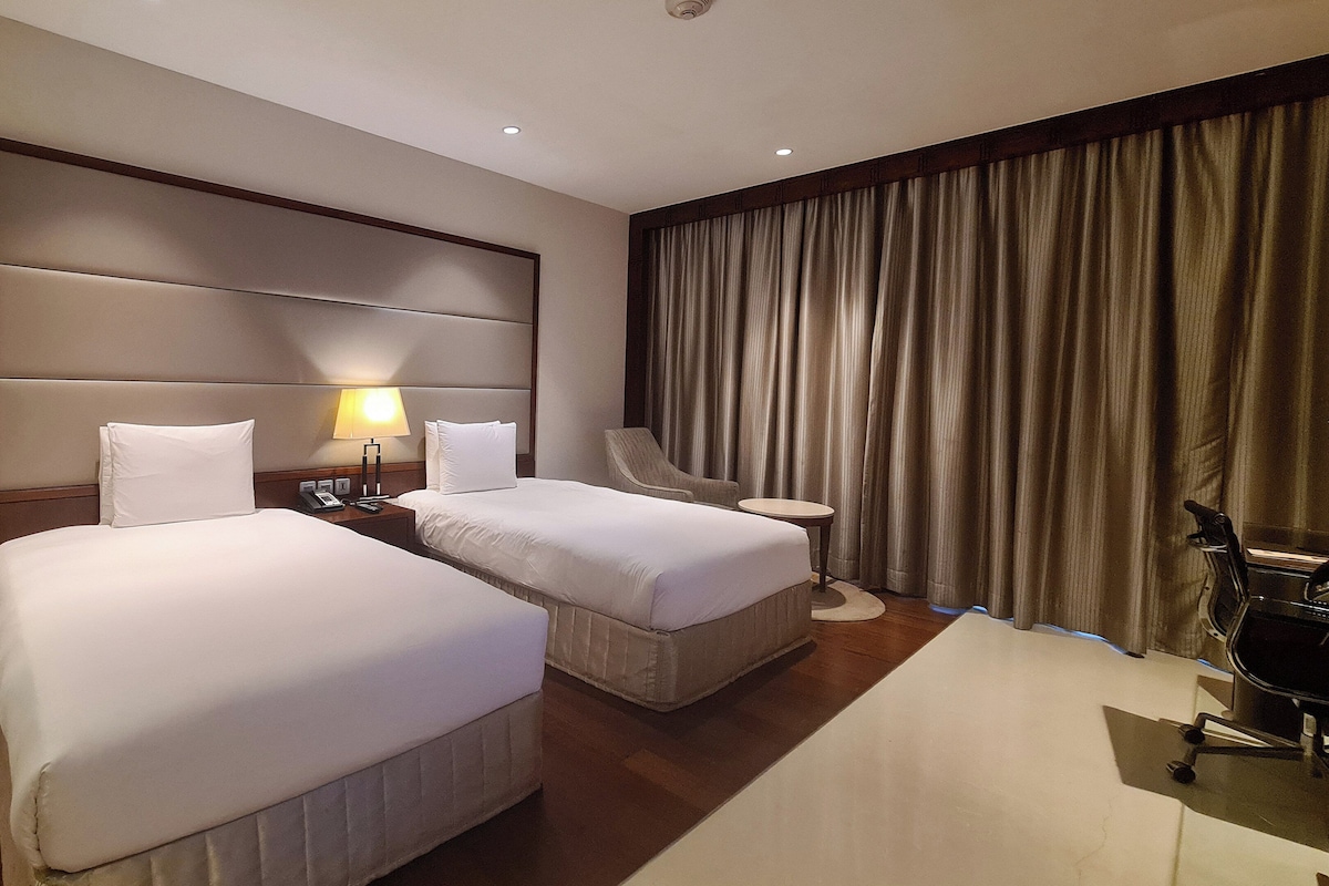 Quarto de hotel com duas camas, mesa de cabeceira, uma cadeira, uma pequena mesa e cortina.
