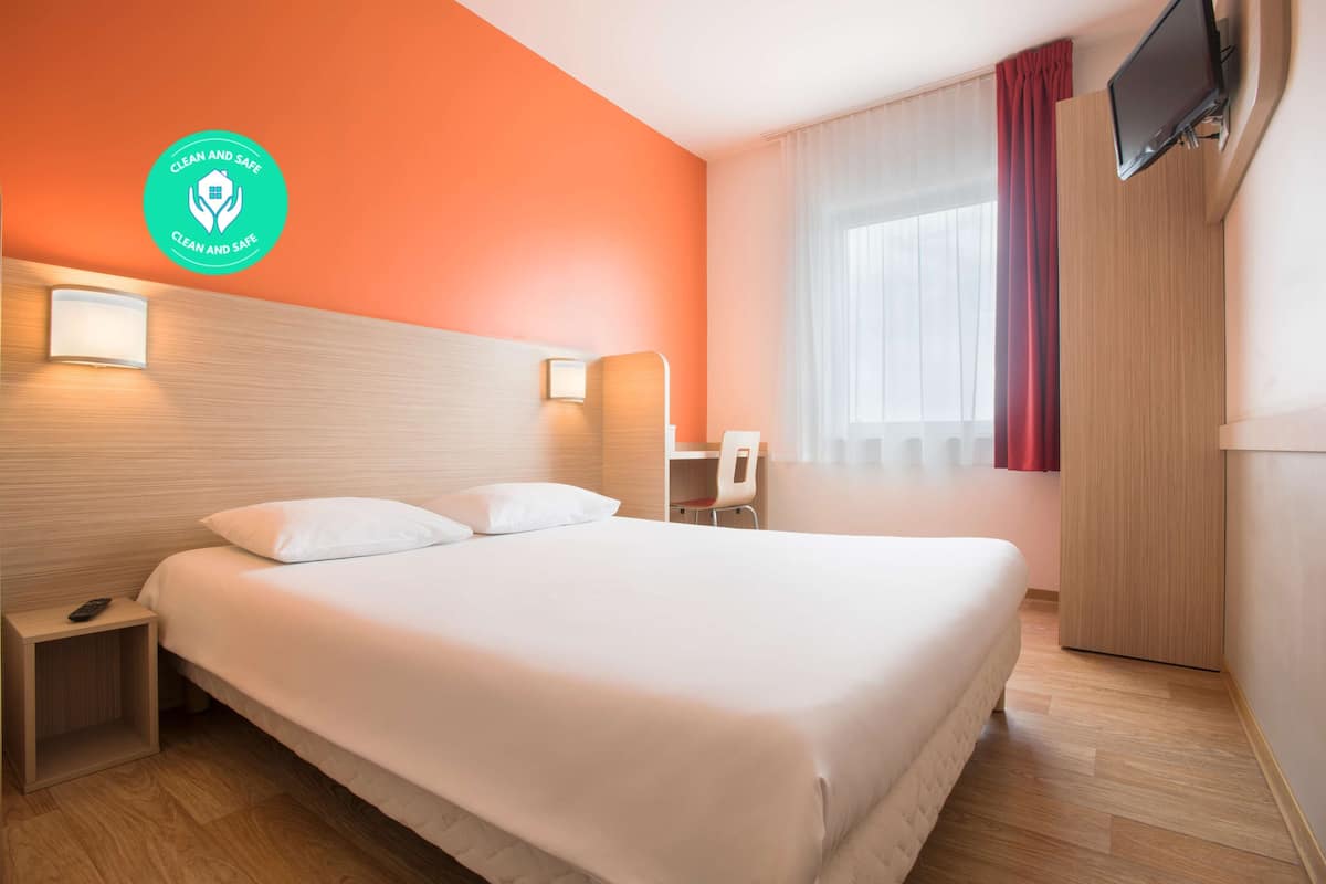 Quarto de hotel com uma cama grande, uma mesa de cabeceira com controle remoto, uma cadeira, uma TV fixada na parede e uma janela com cortinas.