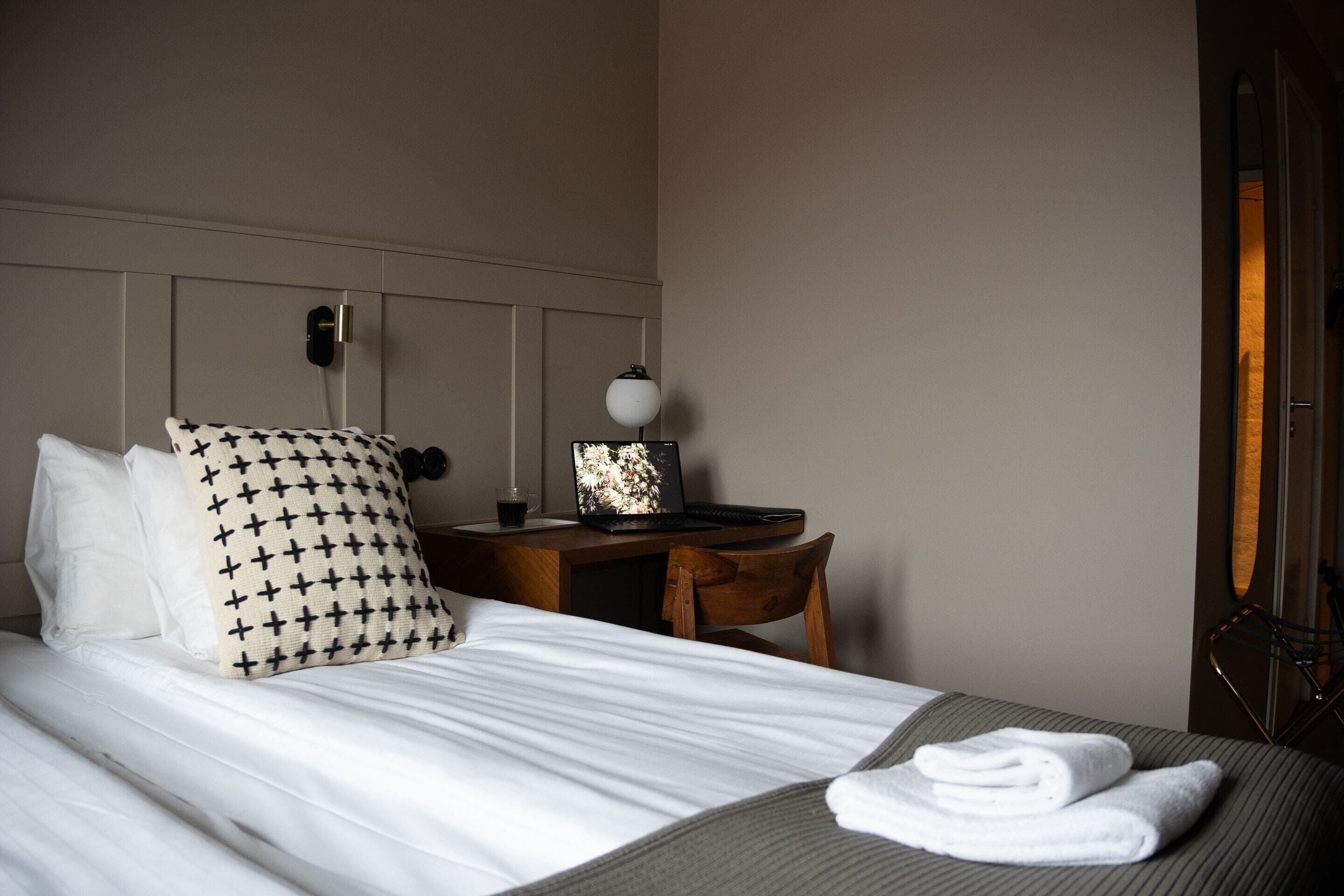Quarto de hotel com cama, escrivaninha com luminária, cadeira e telefone fixado na parede.