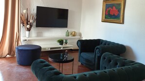 Uma sala de estar com uma televisão de tela plana grande fixada em uma parede branca, dois sofás verde-escuros, uma mesa redonda pequena e um banquinho azul.