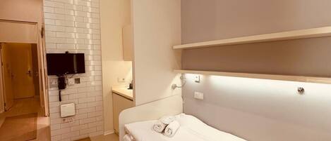 Quarto de hotel moderno com uma cama bem arrumada, uma mesinha de cabeceira e uma TV fixada na parede.