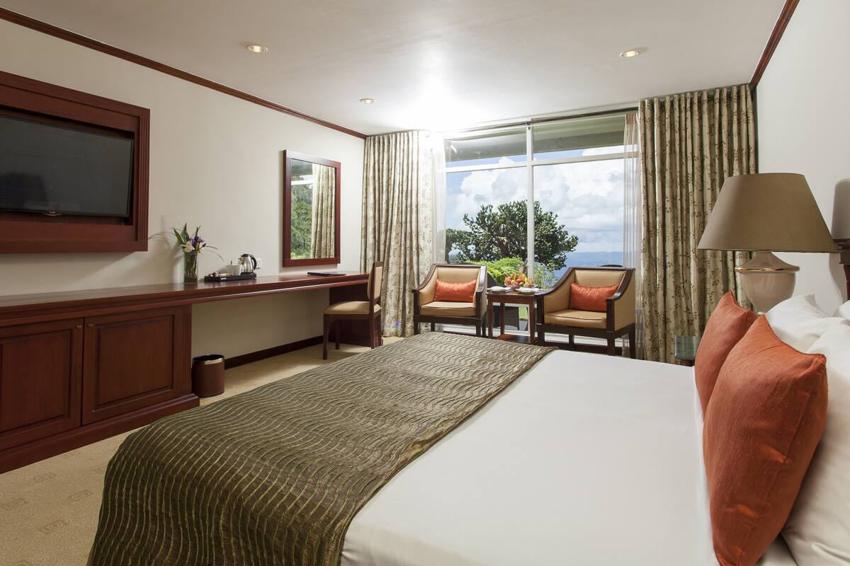 Quarto de hotel com uma cama grande, uma mesa, uma cadeira, uma televisão fixada na parede e uma janela com cortinas.