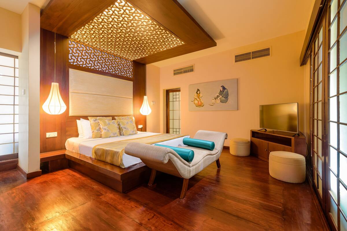 Quarto de hotel com piso de madeira, uma cama com travesseiros, mesa de cabeceira, TV fixada na parede e um painel decorativo no teto.