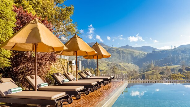 Uma área à beira da piscina com espreguiçadeiras e guarda-sóis, piscina cristalina e montanhas ao fundo.