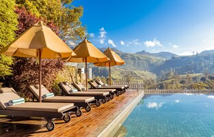 Uma área à beira da piscina com espreguiçadeiras e guarda-sóis, piscina cristalina e montanhas ao fundo.