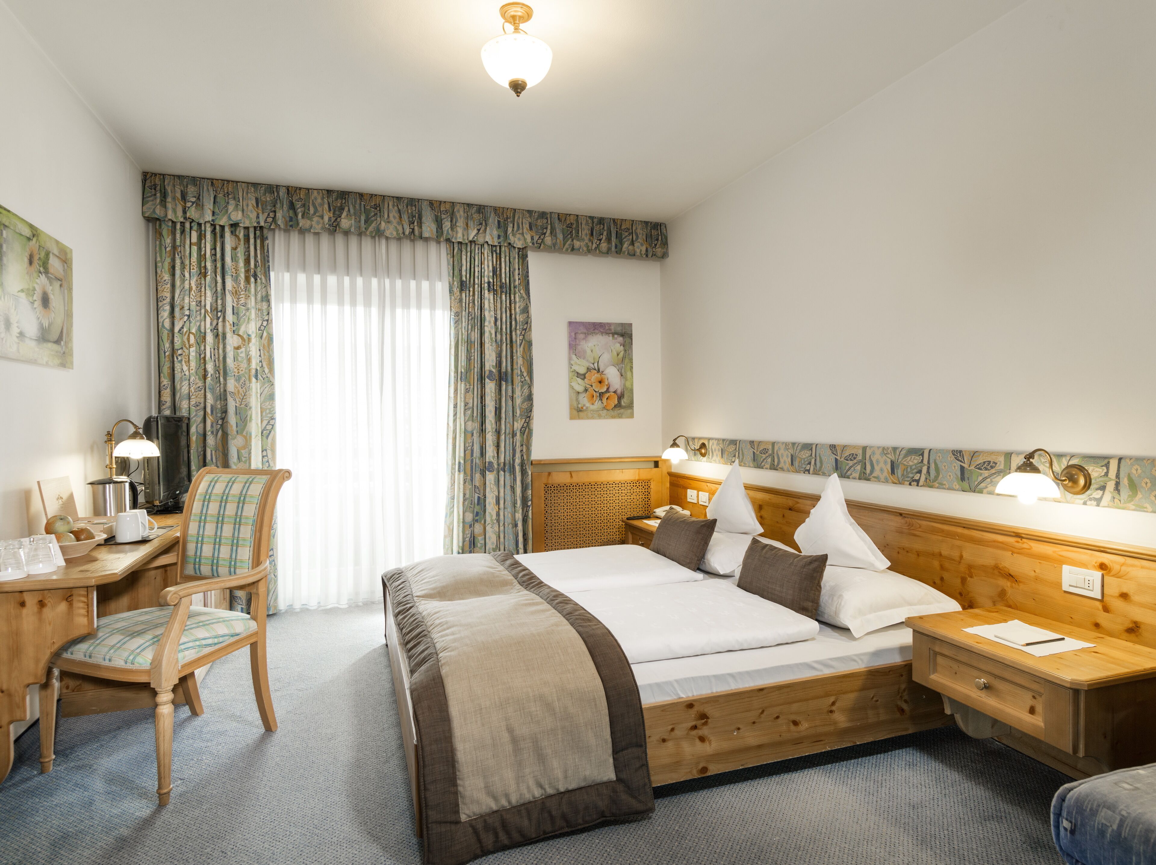 Quarto de hotel com cama de madeira, escrivaninha com luminária, cadeira e janela com cortinas.