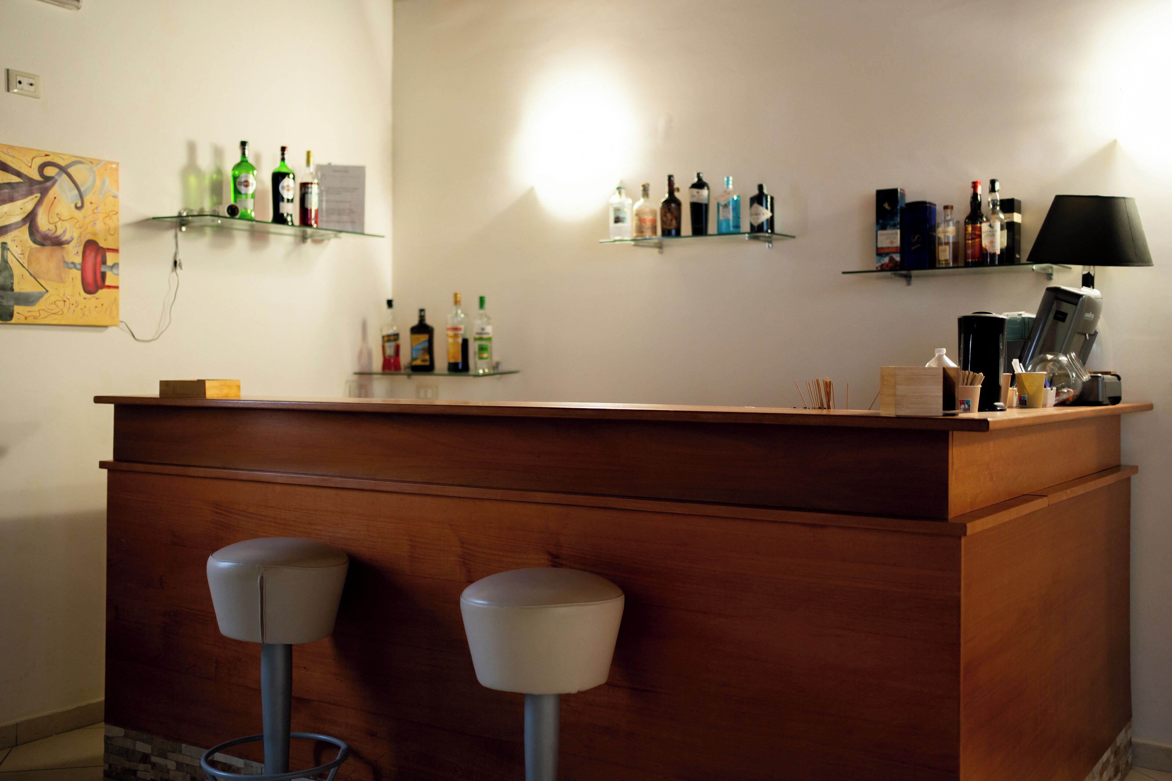 Uma área de bar com banquetas, prateleiras com garrafas, um balcão e um quadro na parede.