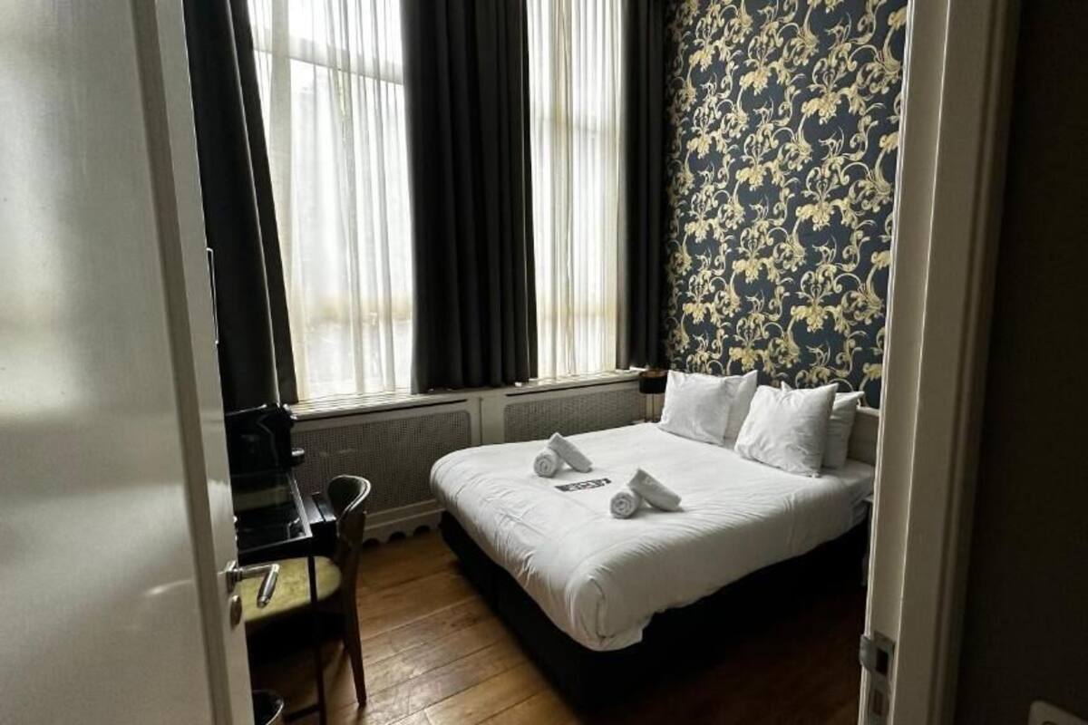 Quarto de hotel com cama, uma cadeira e janela com cortinas.