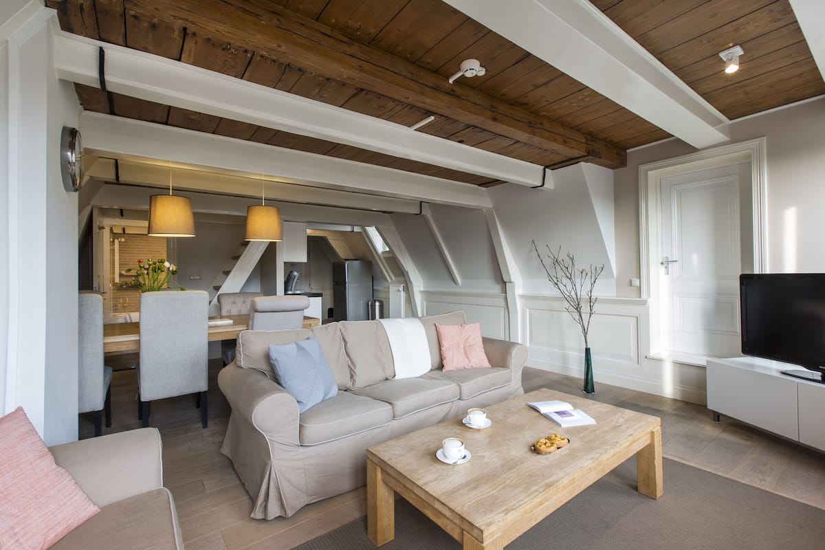 Uma sala de estar moderna com sofá, mesa de centro e televisão. Há uma área de jantar com cadeiras e mesa ao fundo.