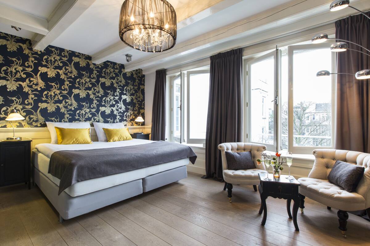Quarto com uma cama grande, duas poltronas, uma mesinha de cabeceira, um candelabro e vista da cidade por grandes janelas.