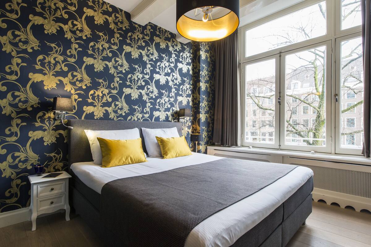 Quarto com cama grande, mesa de cabeceira, luminária e janela com vista para prédios.