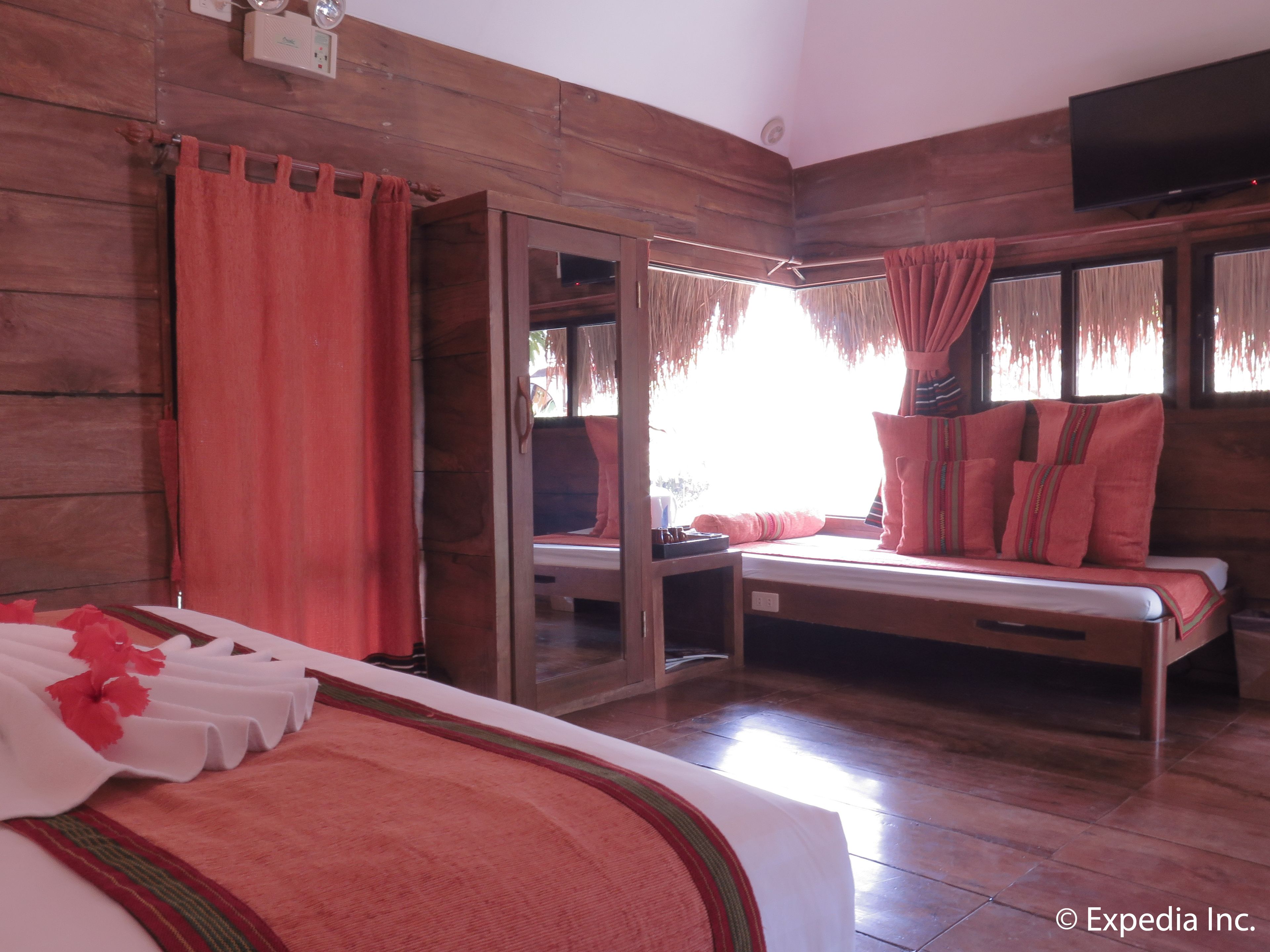 Quarto com paredes de madeira, cama com manta vermelha, televisão na parede e janela com cortinas.