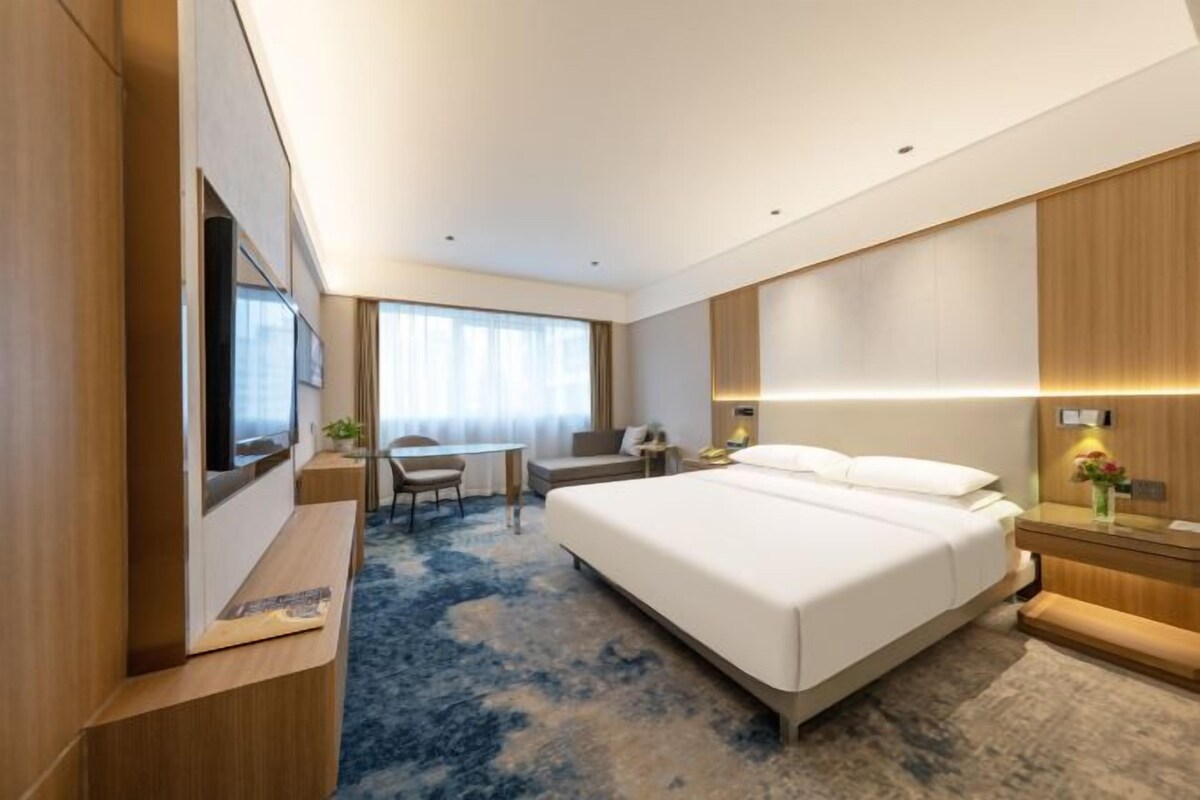 Quarto de hotel moderno com uma cama grande, uma escrivaninha com cadeira, televisão de tela plana e uma janela ampla com cortinas.