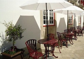 Área de pátio externa com cadeiras e mesas de metal vermelho, guarda-sóis brancos e um vaso de planta.