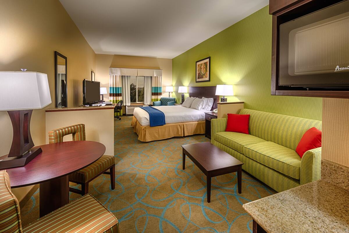 Quarto de hotel com cama, sofá verde, mesa de jantar com cadeiras, televisão e escrivaninha com luminária.