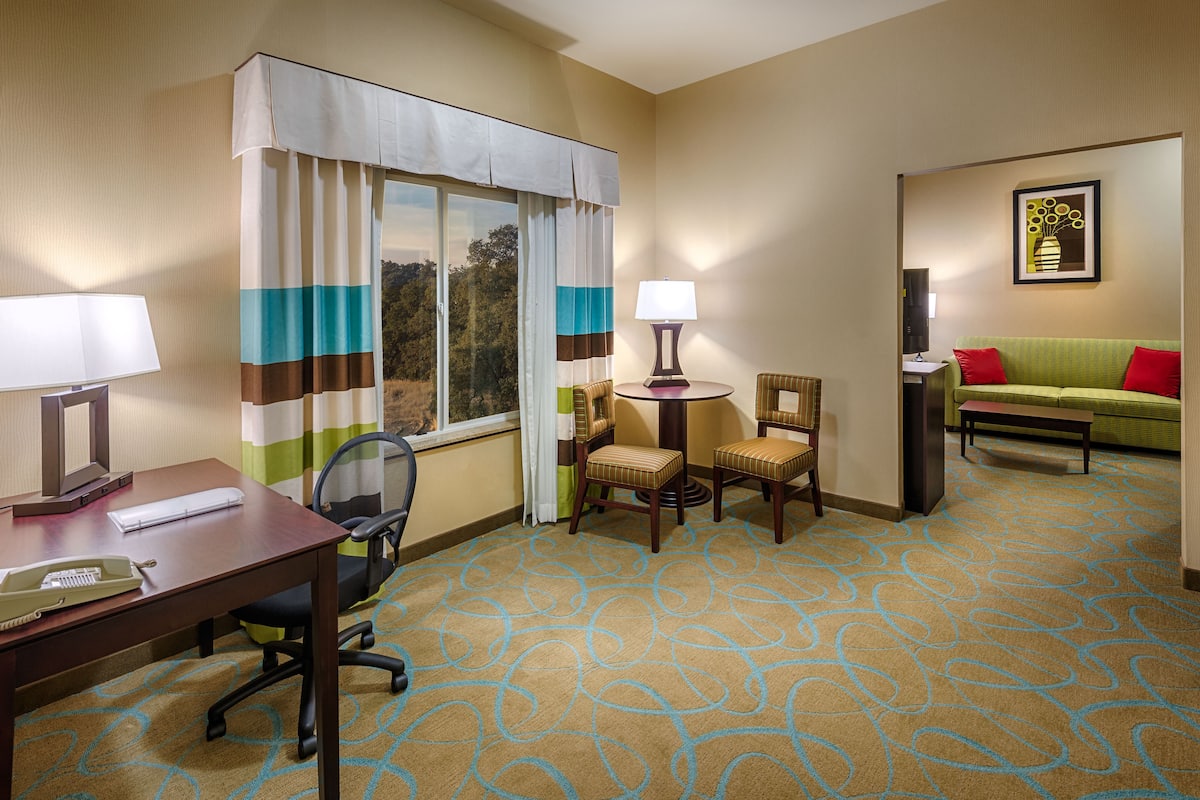 Quarto de hotel com escrivaninha, duas cadeiras, mesa redonda com abajur, sofá verde com almofadas vermelhas e um quadro na parede.