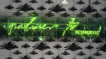 Um letreiro de neon com as palavras 'nature' e 'restaurant' numa parede de vidro refletora com padrões de palmeiras.