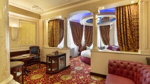 Quarto de hotel luxuoso com tapete vermelho, um espelho grande, sofá vermelho com detalhes em capitonê e uma cama com cabeceira branca.