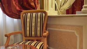 Uma cadeira de madeira com almofada listrada, uma mesa redonda de madeira com tampo de vidro e uma janela com cortinas vermelhas e creme.