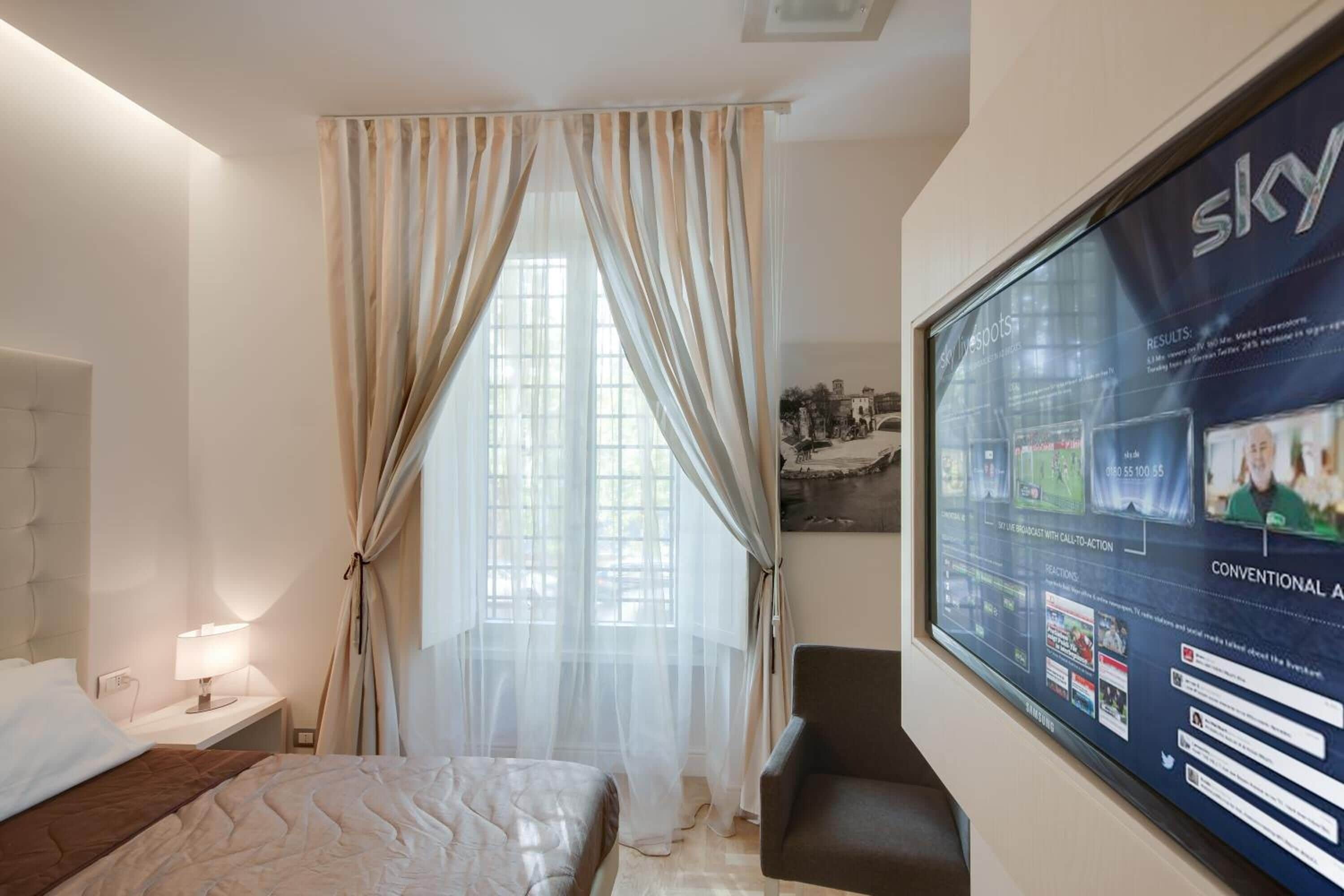 Quarto de hotel com cama, escrivaninha com luminária, uma cadeira e uma tela grande exibindo informações.
