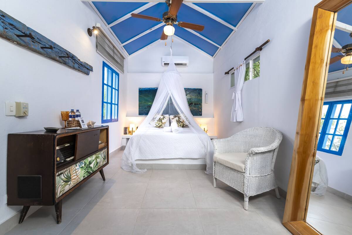 Quarto com cama de dossel, armário de madeira, cadeira de vime e ventilador de teto.